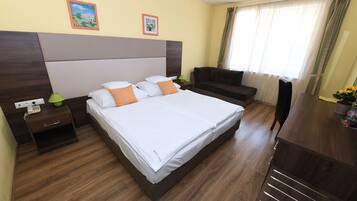 Quarto de hotel com uma cama grande, mesa de cabeceira com abajur, pequena escrivaninha, sofá e janela com cortinas.