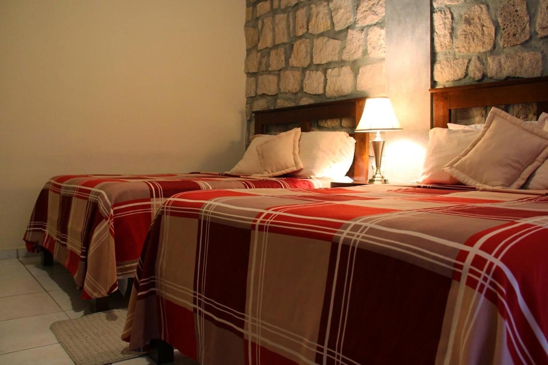 Quarto de hotel com duas camas, cada uma com colcha xadrez vermelha e bege, uma parede de pedra e uma luminária de cabeceira.