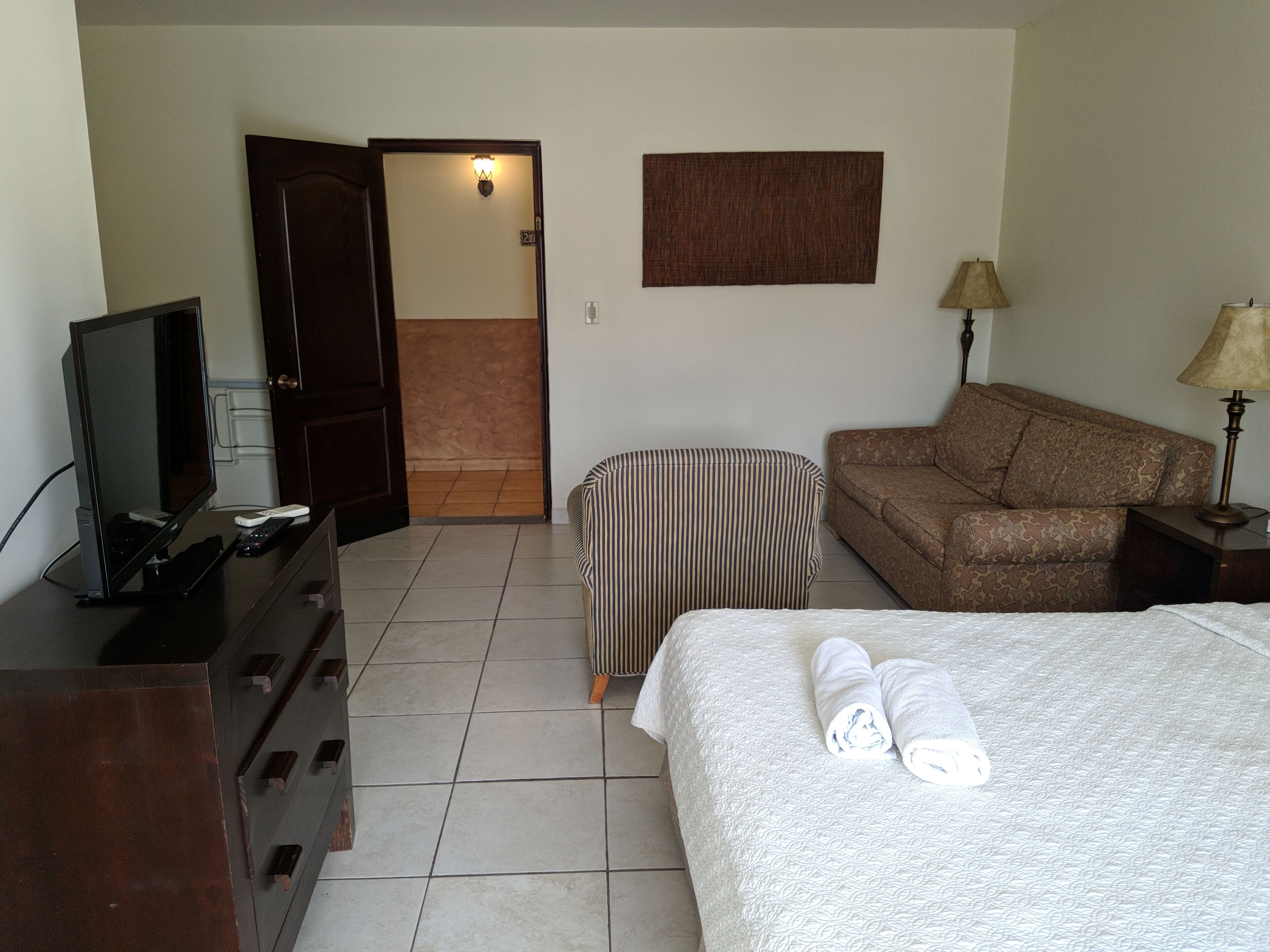 Quarto de hotel com uma cama, um sofá, uma televisão sobre uma cômoda e uma porta que dá para o banheiro.