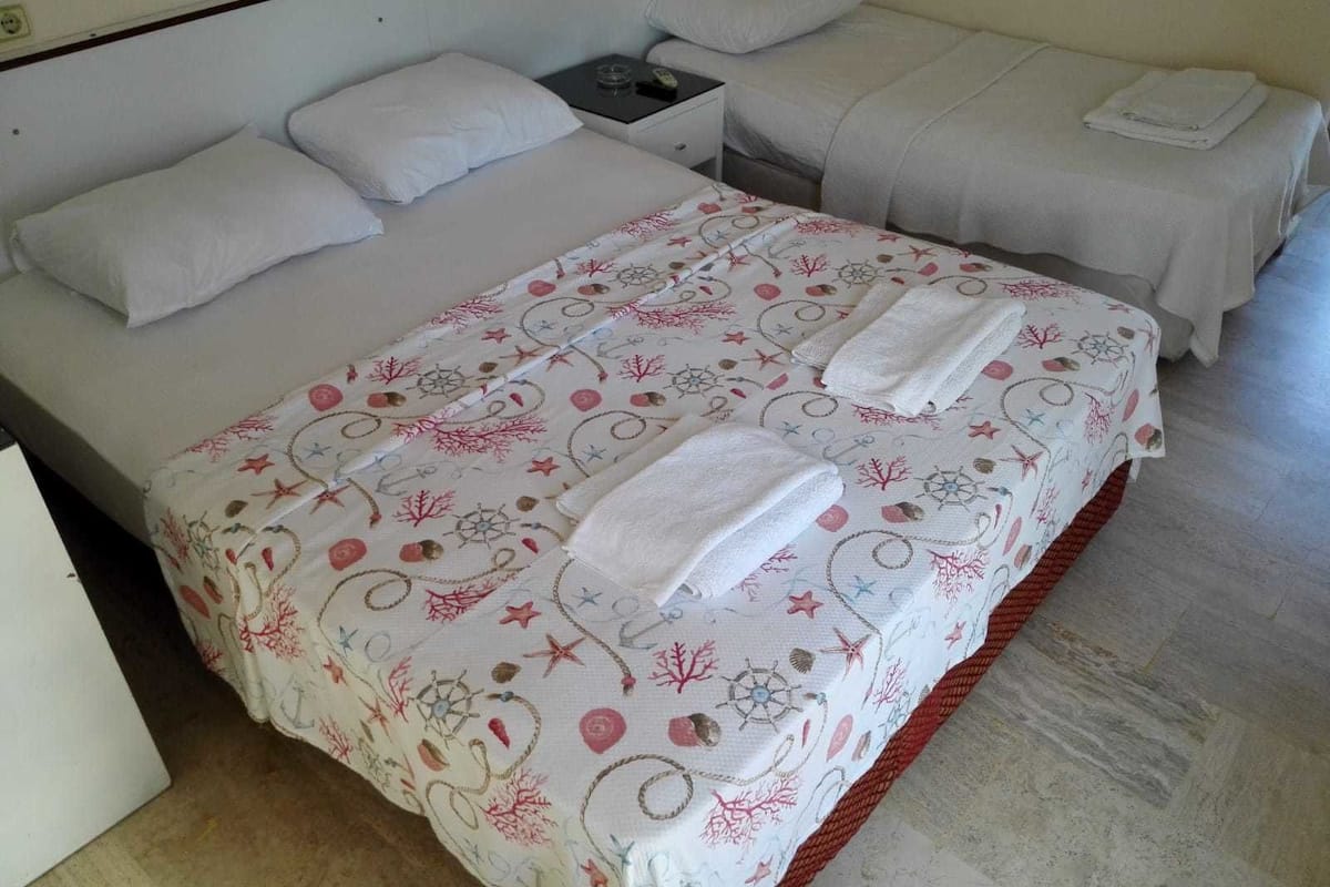Uma cama com colcha estampada, duas almofadas, uma mesa de cabeceira com abajur e um criado-mudo pequeno.