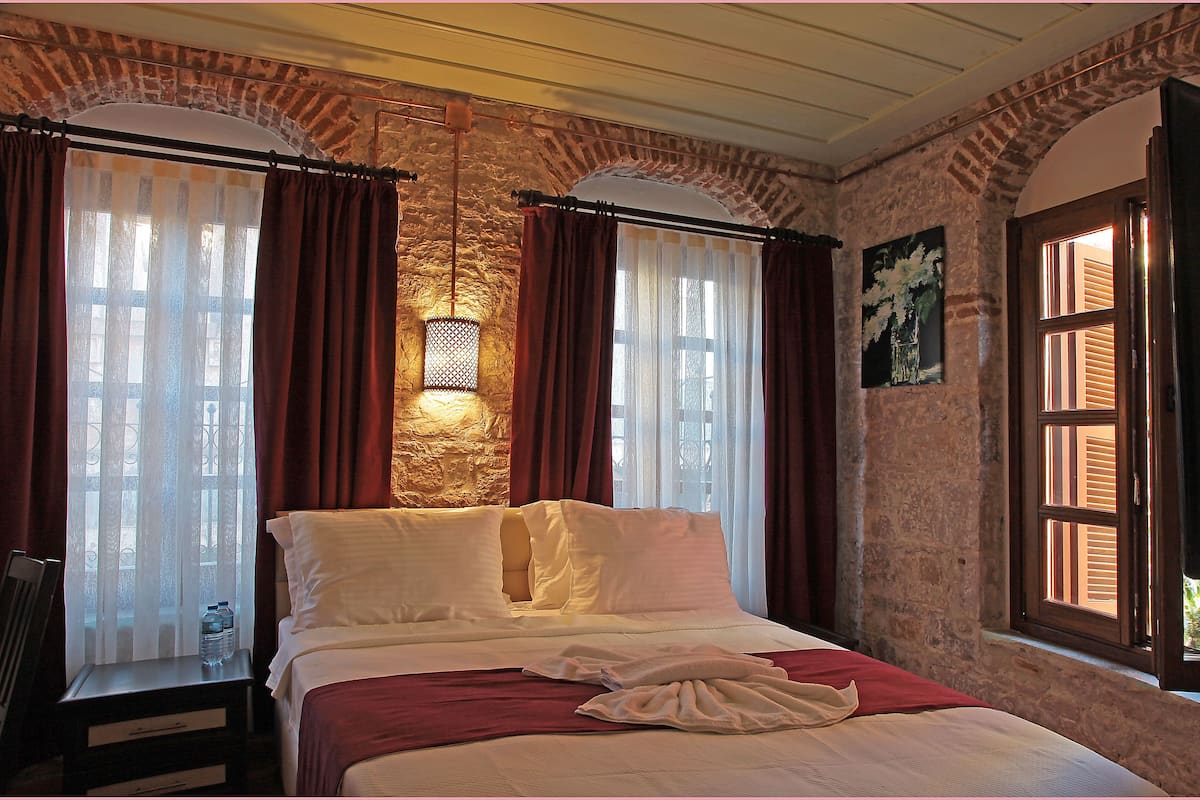 Quarto de hotel com uma cama grande, duas janelas com cortinas, uma mesinha com uma cadeira e uma parede de pedra.