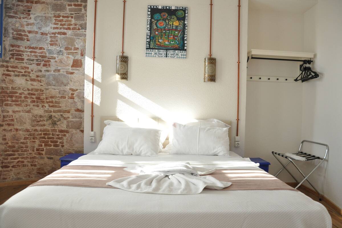 Cama bem arrumada com lençóis brancos, uma obra de arte pendurada e uma parede de tijolos.