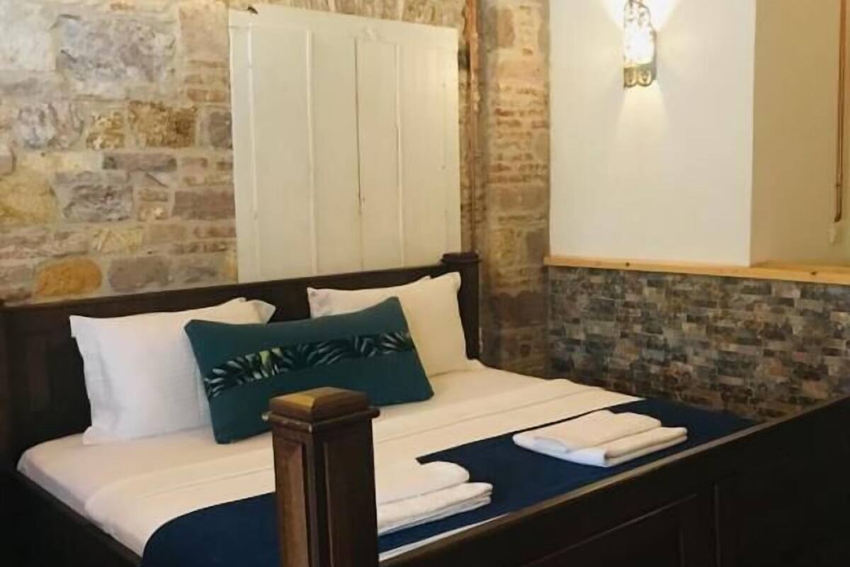 Quarto com parede de pedra, cama de madeira com almofadas brancas e azul-turquesa, guarda-roupa branco e espelho com luminária.