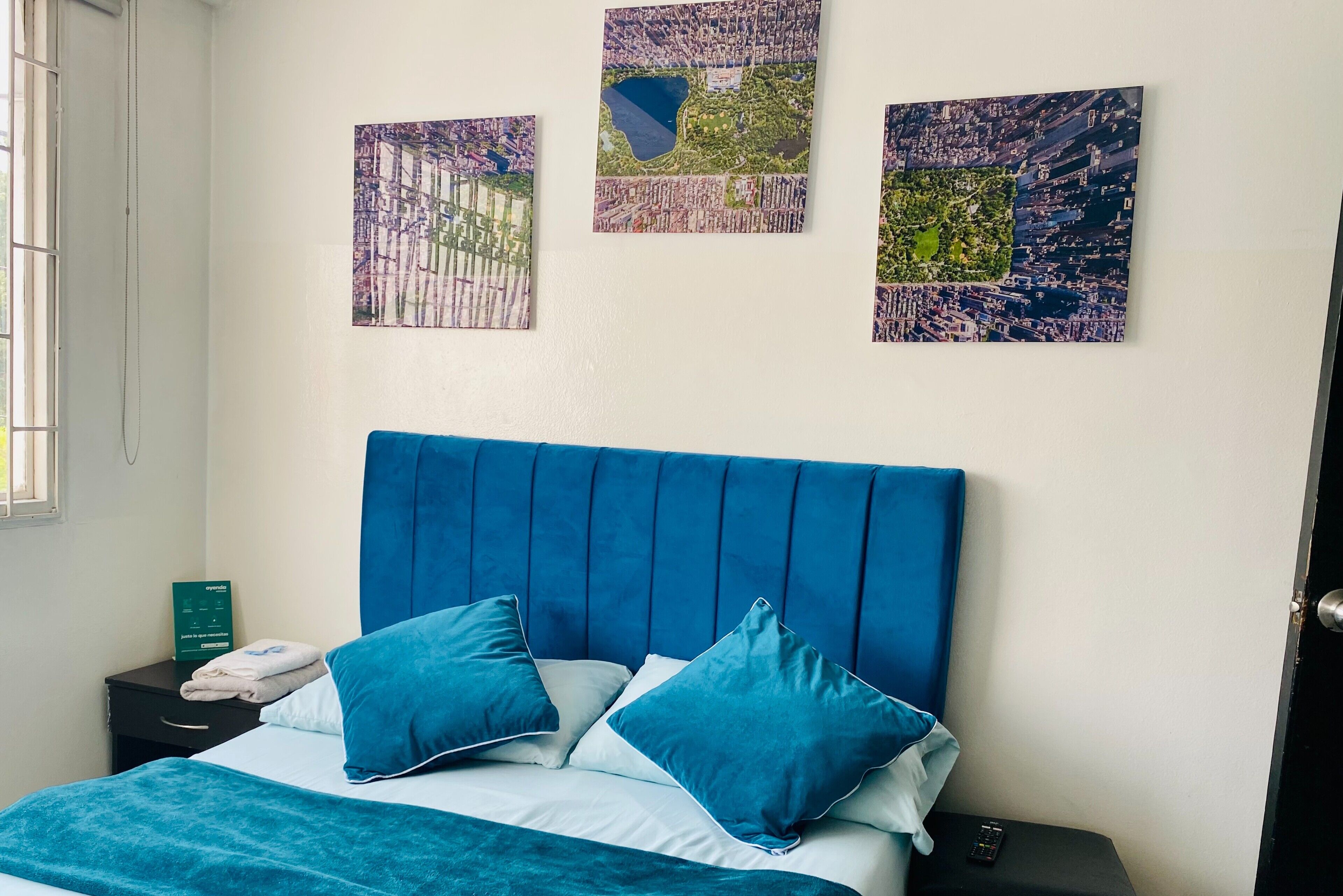 Quarto com cabeceira azul, cama com roupa de cama azul e branca e três fotografias aéreas emolduradas de paisagens urbanas na parede.