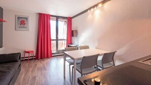 Quarto compacto com mesa de jantar, cadeiras, sofá, televisão e janela com cortinas vermelhas.