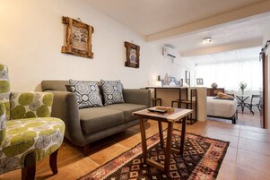 Uma sala de estar com um sofá, uma mesa de centro e um tapete estampado. Ao fundo, há uma área de jantar com mesa e cadeiras.