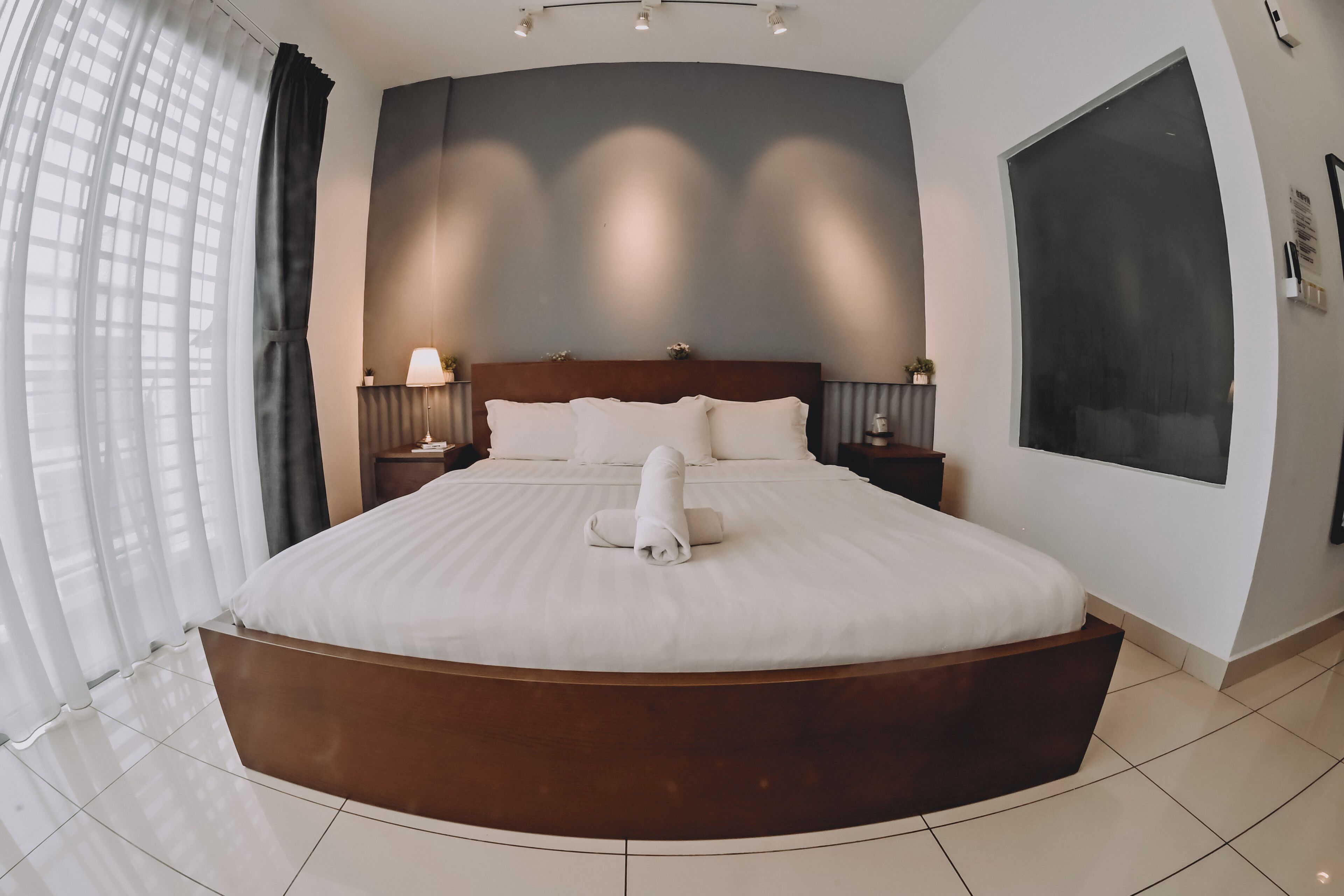 Quarto de hotel moderno com cama curva, uma janela grande com cortinas finas e vista para a paisagem urbana.