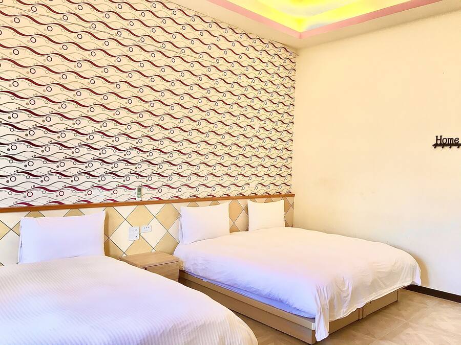 Quarto de hotel com duas camas, papel de parede estampado e um letreiro com a palavra 'Home'.