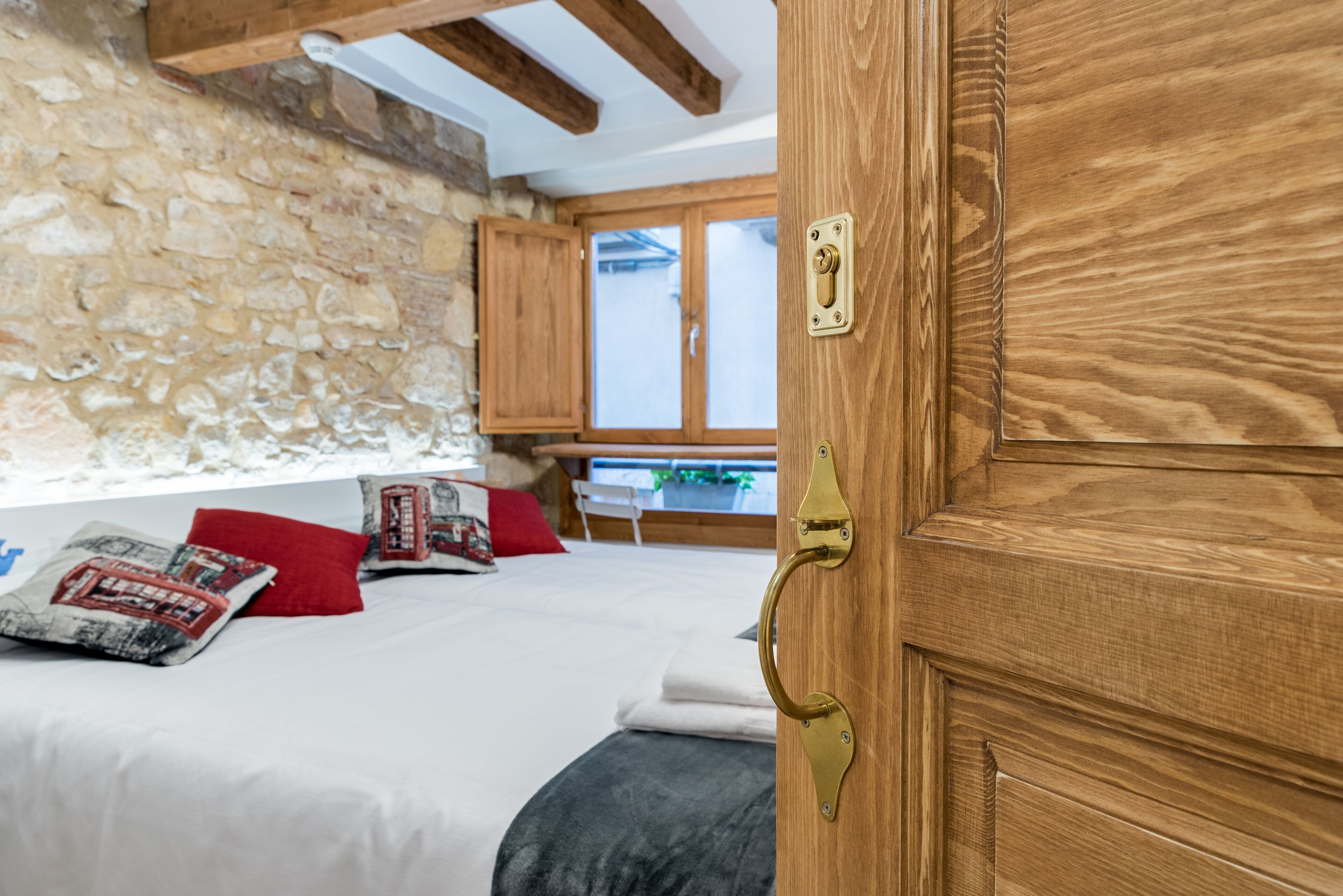 Quarto com parede de pedra, vigas de madeira no teto, cama com roupa de cama branca e almofadas vermelhas, e porta de madeira com puxador de latão.