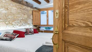 Quarto com parede de pedra, vigas de madeira no teto, cama com roupa de cama branca e almofadas vermelhas, e porta de madeira com puxador de latão.