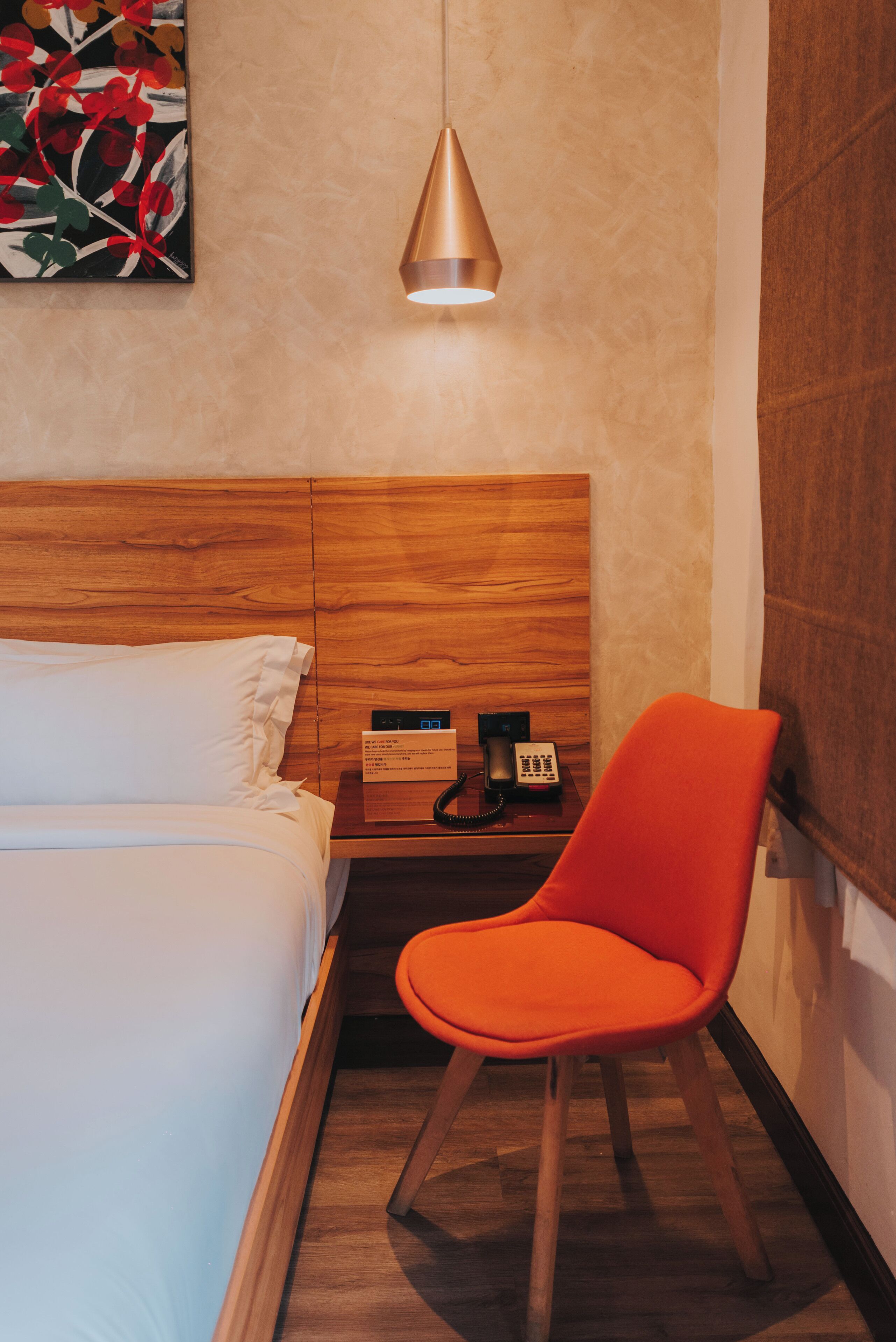Quarto de hotel com cabeceira de madeira, cama com roupa de cama branca, uma cadeira laranja, mesa de cabeceira com telefone e uma caixa pequena, e luminária pendente.