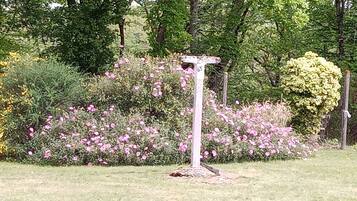 Um jardim com uma estrutura em forma de cruz branca, rodeado por diversas plantas floridas e árvores.