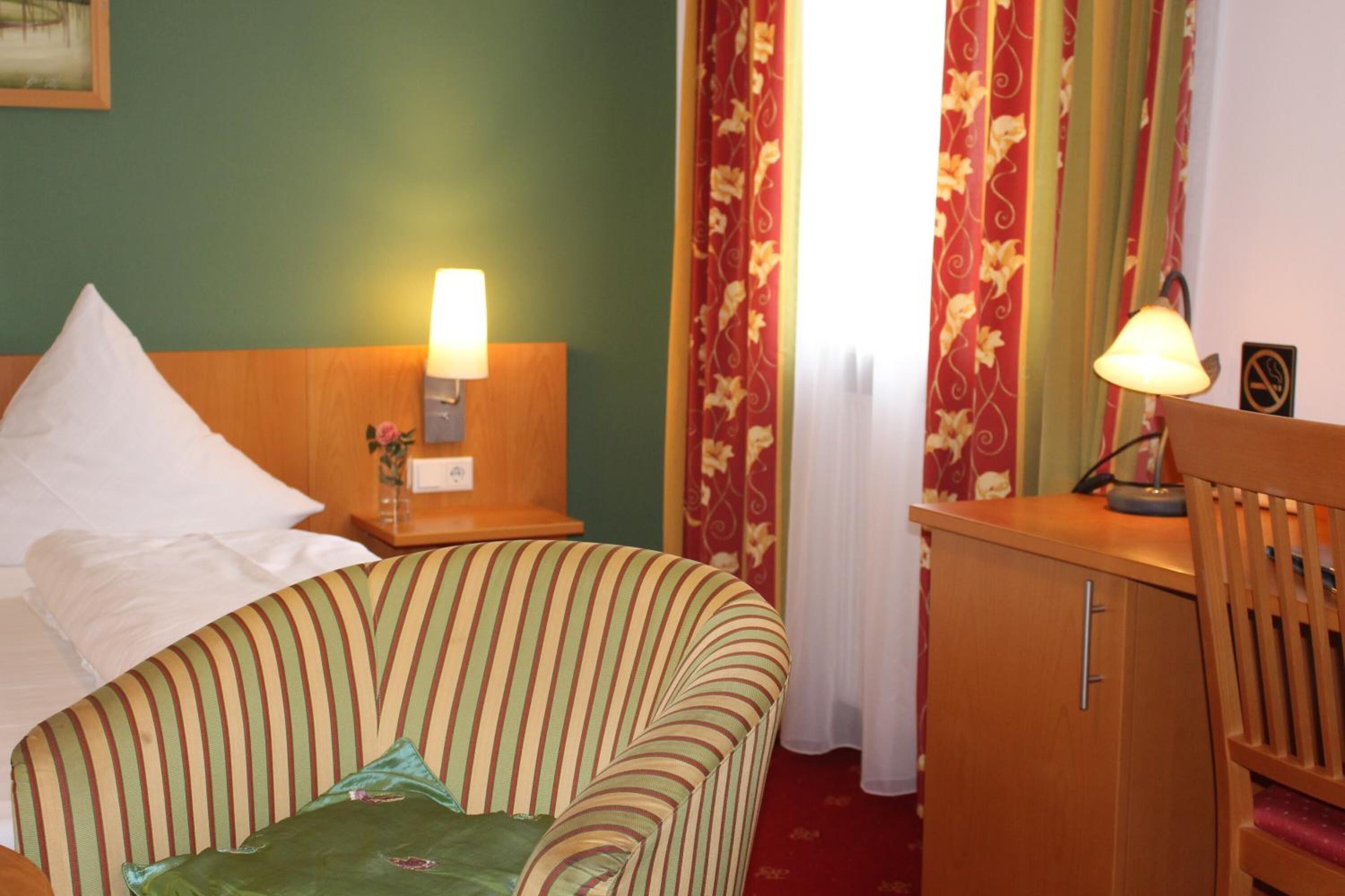 Quarto de hotel com uma sofá-cama listrada, uma mesinha de cabeceira de madeira com abajur, um pequeno armário e uma janela com cortinas vermelhas.