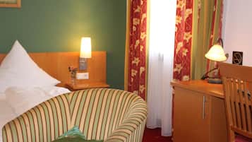 Quarto de hotel com uma sofá-cama listrada, uma mesinha de cabeceira de madeira com abajur, um pequeno armário e uma janela com cortinas vermelhas.