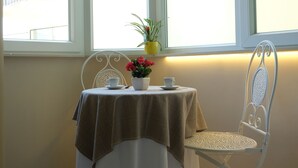 Quarto com mesa posta para dois, uma cadeira, um vaso de planta e vista para um edifício.