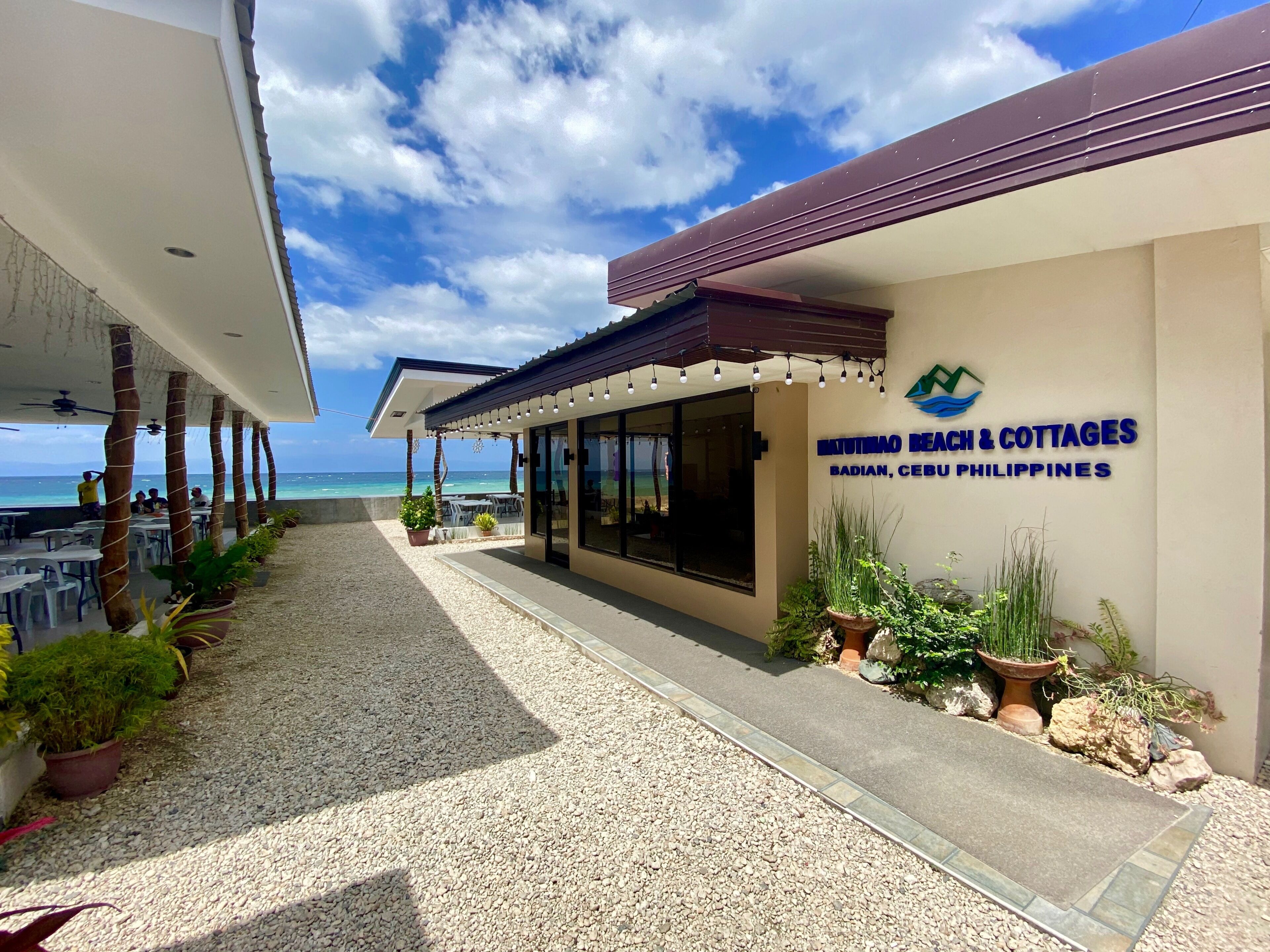 Uma área externa coberta com mesas e cadeiras, um edifício com a placa 'KUMIMO BEACH & COTTAGES' e vista para o oceano.