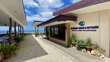 Uma área externa coberta com mesas e cadeiras, um edifício com a placa 'KUMIMO BEACH & COTTAGES' e vista para o oceano.