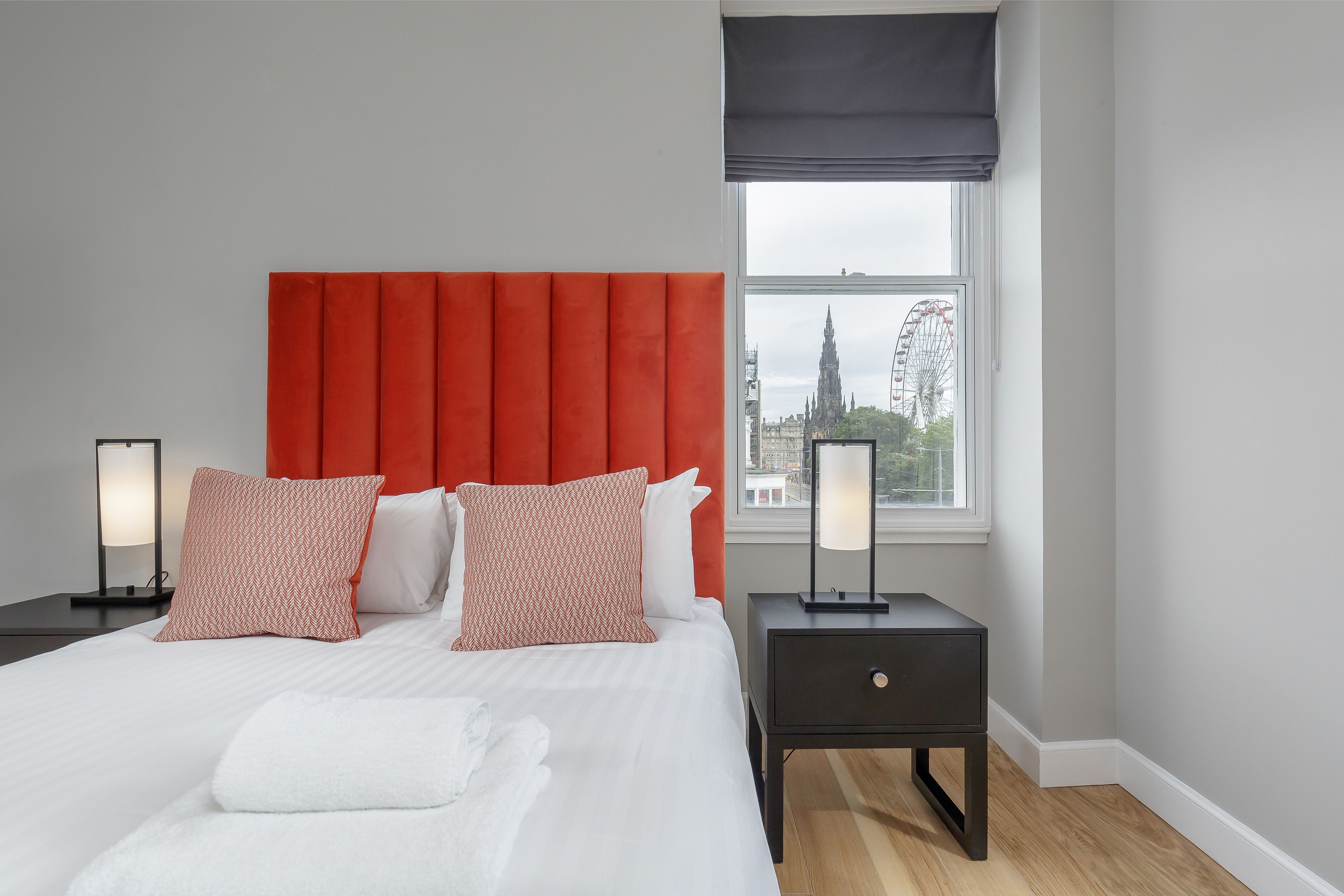 Quarto de hotel com cama, duas mesinhas de cabeceira, janela com vista para a paisagem urbana e cabeceira vermelha.