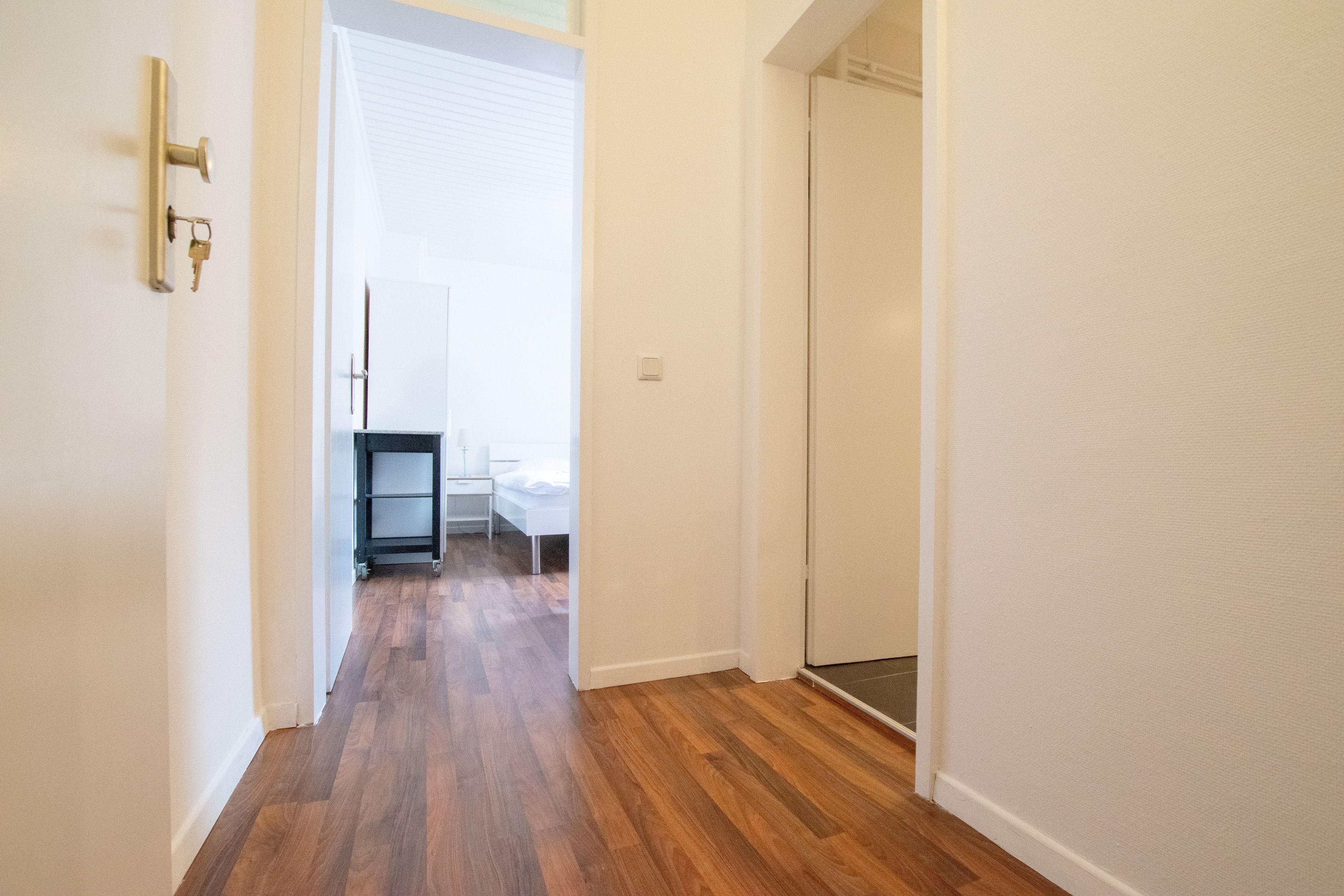 Um corredor com piso de madeira, uma porta com fechadura e uma visão de um quarto com cama e janela.