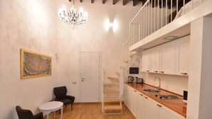Uma cozinha compacta com armários brancos, bancada de madeira e pia embutida. Há uma pequena área de jantar com duas cadeiras e uma mesa, e uma escada que leva a um andar superior.
