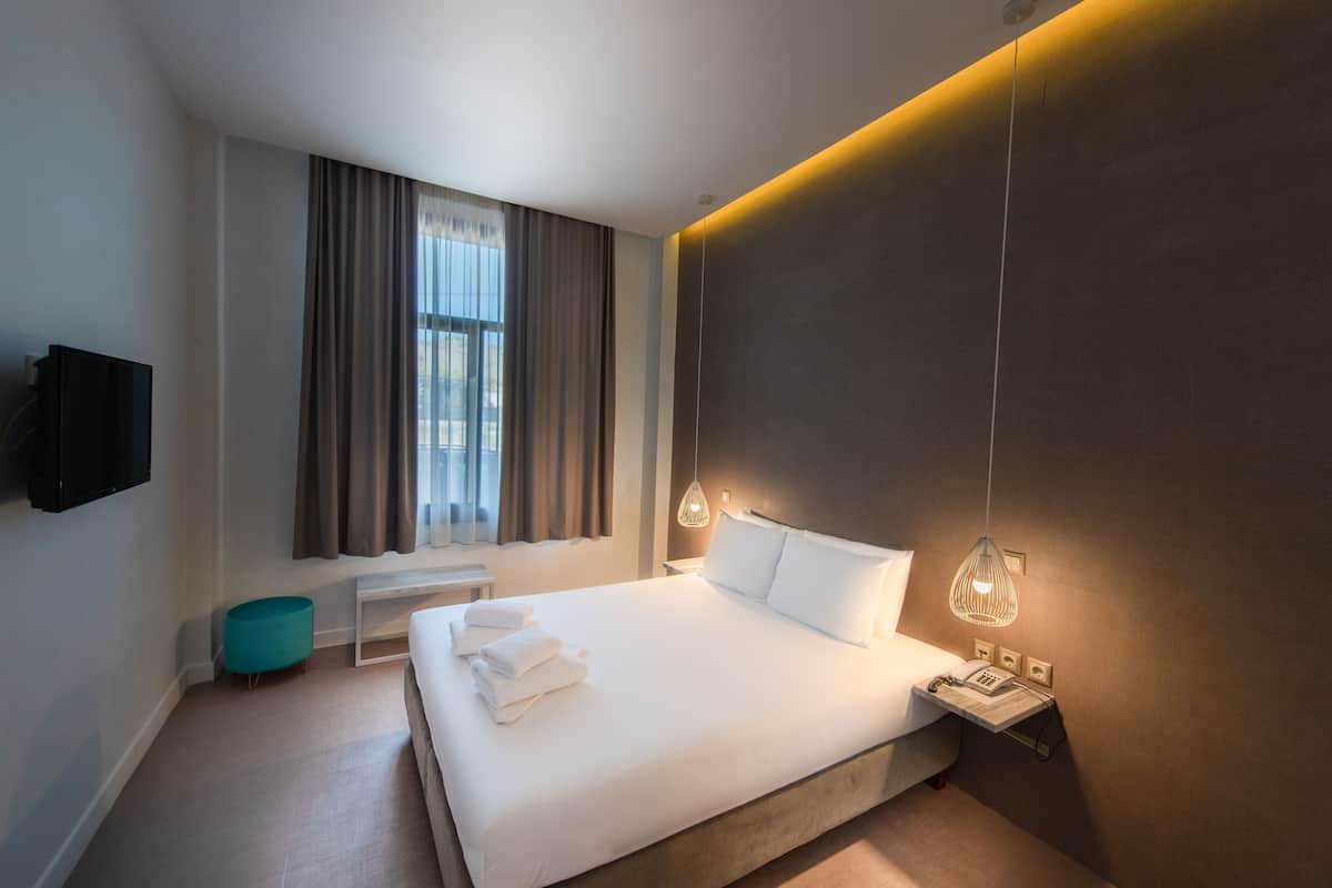 Quarto de hotel moderno com uma cama grande, televisão de tela plana na parede, mesa de cabeceira com abajur e uma janela com cortinas.