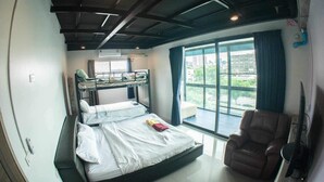 Quarto com beliches, uma cama de solteiro, uma cadeira e uma janela grande com vista para prédios.