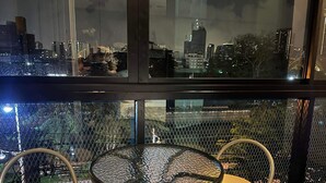 Uma varanda com mesa de vidro e duas cadeiras, com vista para um horizonte urbano à noite.