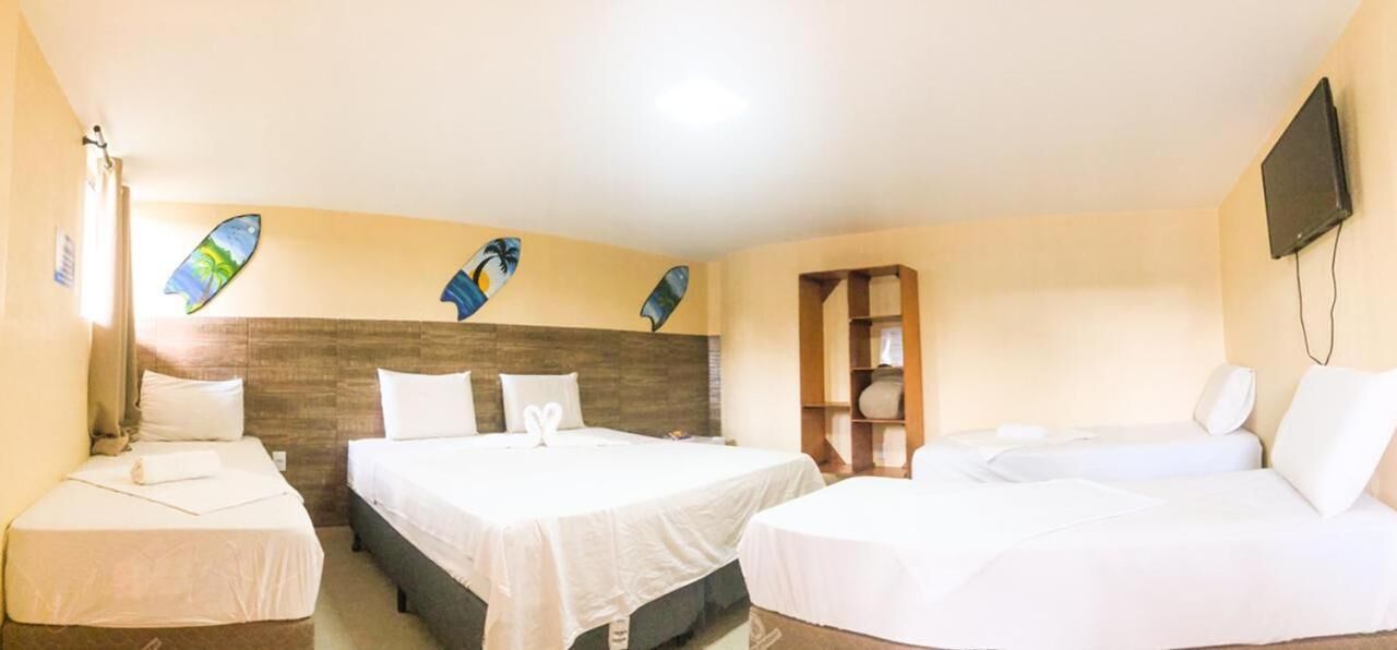 Quarto de hotel com duas camas, cabeceira de madeira, televisão e prateleira com toalhas.