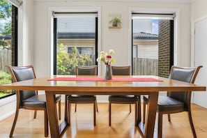 Uma sala de jantar com mesa e cadeiras de madeira, um vaso com flores e vista através de portas deslizantes abertas.