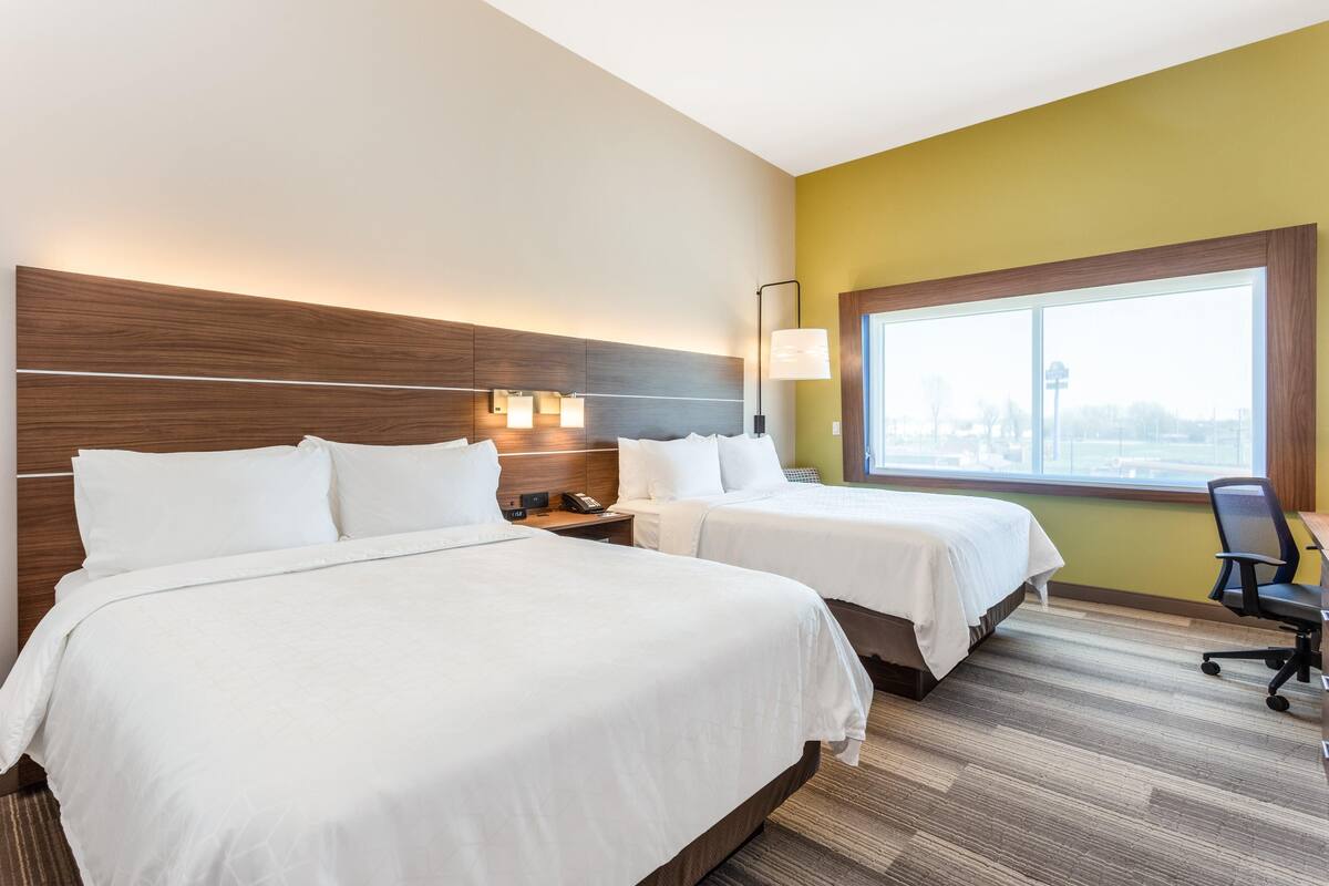 Quarto de hotel com duas camas, uma escrivaninha, uma cadeira, mesa de cabeceira e janela com vista.