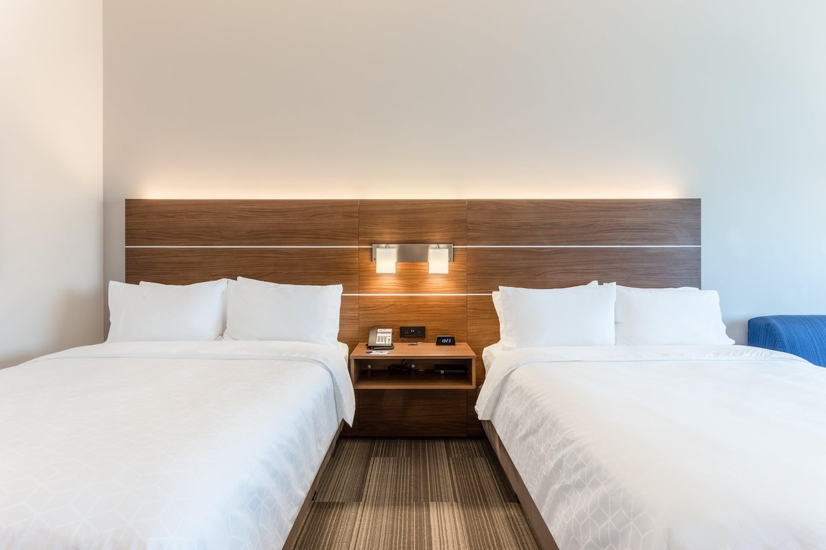 Quarto de hotel com duas camas, cabeceira de madeira, mesas de cabeceira com abajures e um criado-mudo com telefone e luminária.