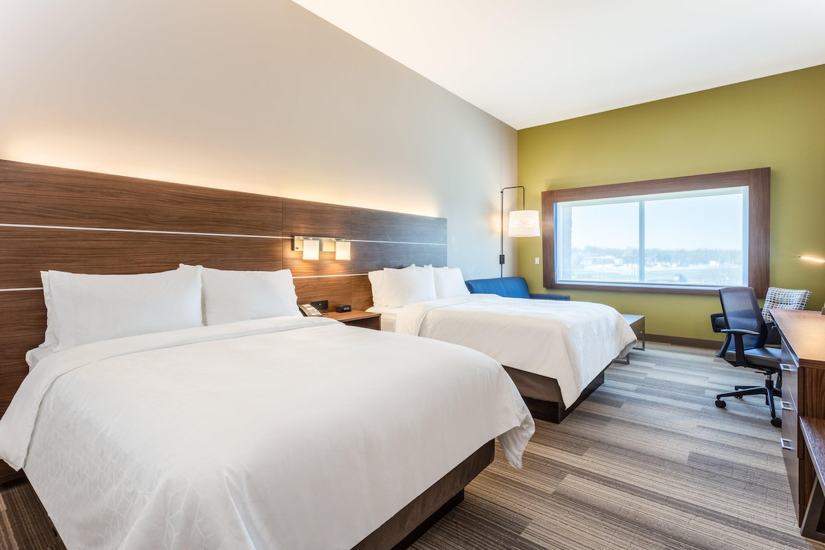 Quarto de hotel com duas camas, uma mesa, uma cadeira, uma luminária e uma janela com vista.
