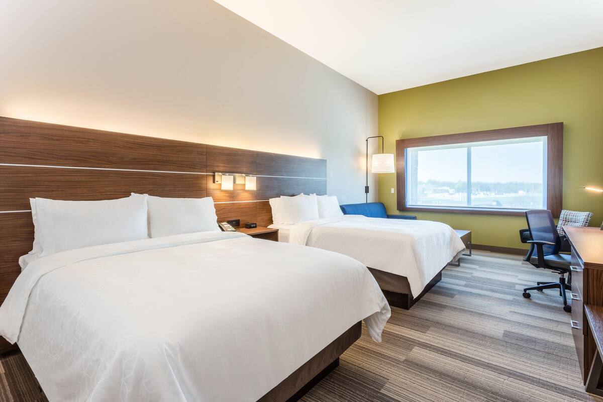 Quarto de hotel com duas camas, uma mesa, uma cadeira, uma luminária e uma janela com vista.