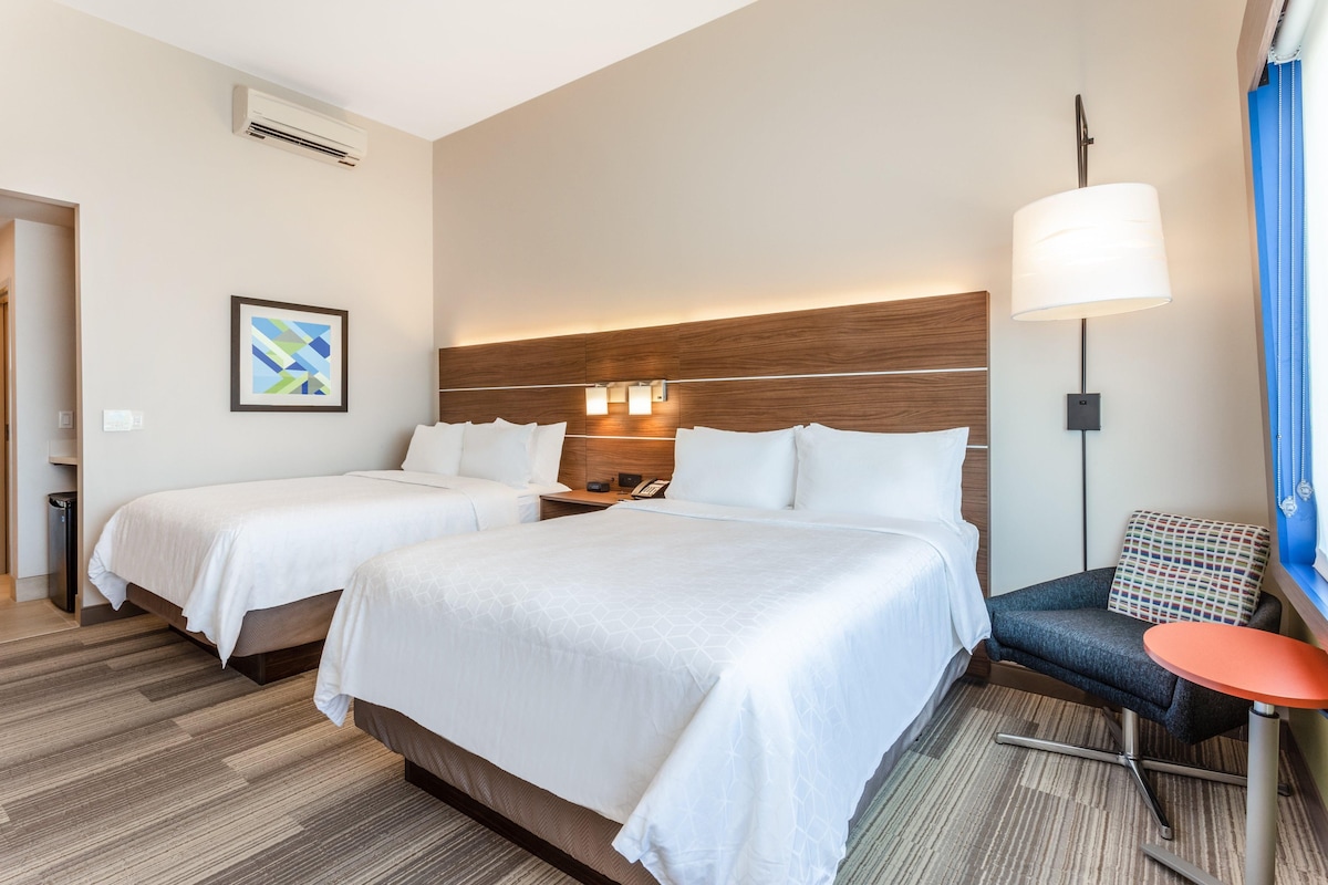 Quarto de hotel com duas camas, uma escrivaninha, uma cadeira, um abajur e um quadro na parede.