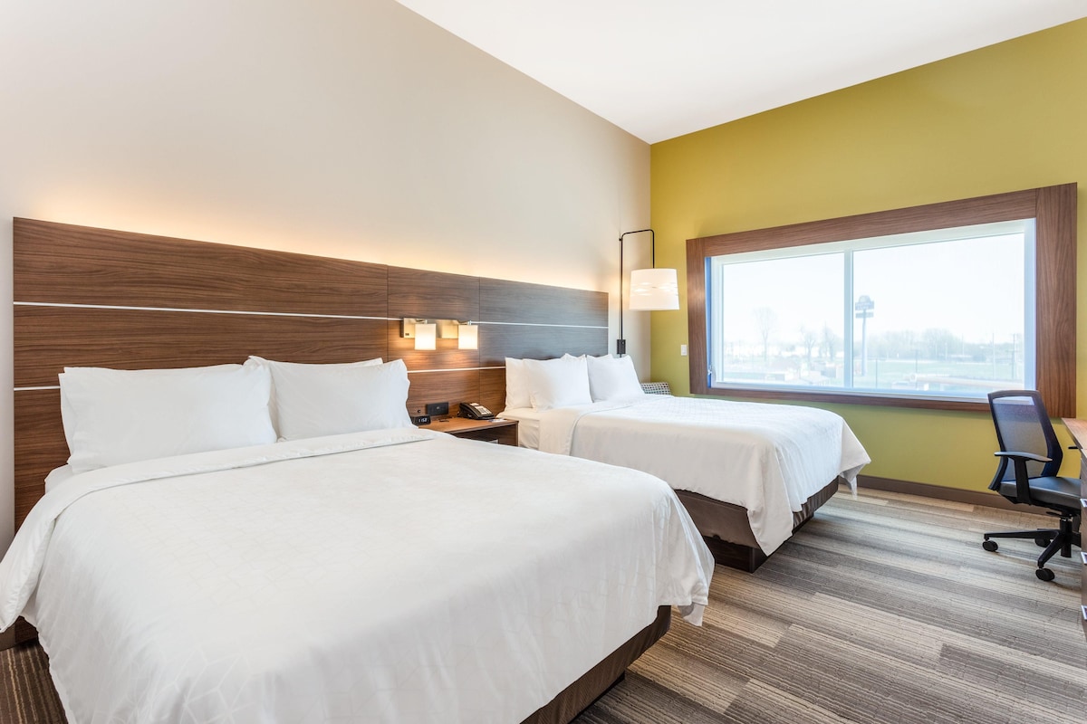 Quarto de hotel com duas camas, uma escrivaninha, uma cadeira, mesa de cabeceira e janela com vista.