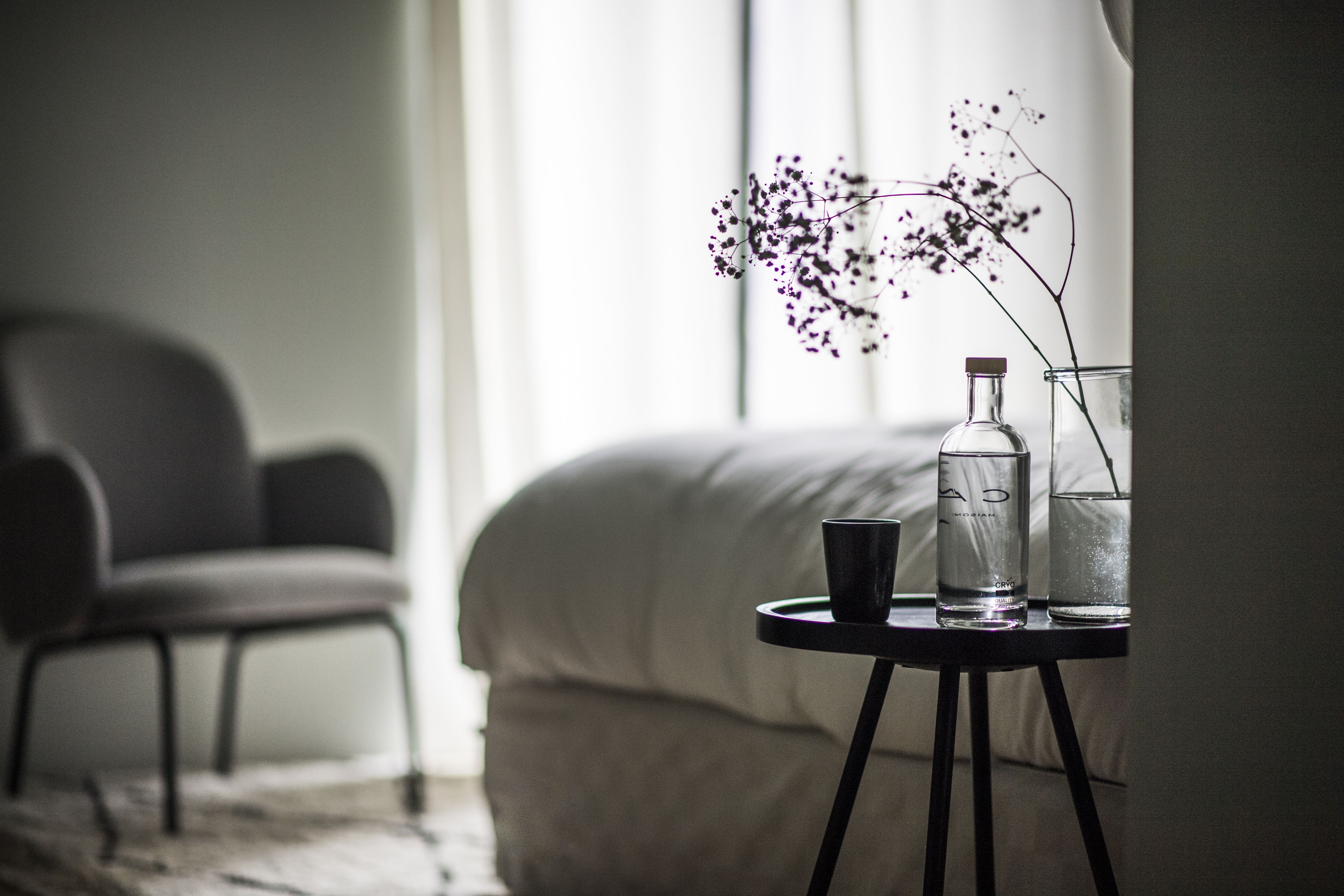 Quarto com cama, uma cadeira, uma mesa pequena com uma garrafa e uma xícara, e um vaso com flores.