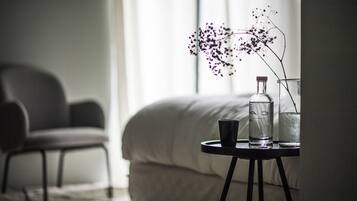 Quarto com cama, uma cadeira, uma mesa pequena com uma garrafa e uma xícara, e um vaso com flores.