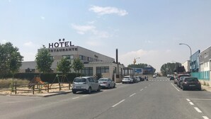 Um hotel com uma placa que diz "HOTEL RESTAURANTE" numa rua ensolarada, com carros estacionados e um parquinho.
