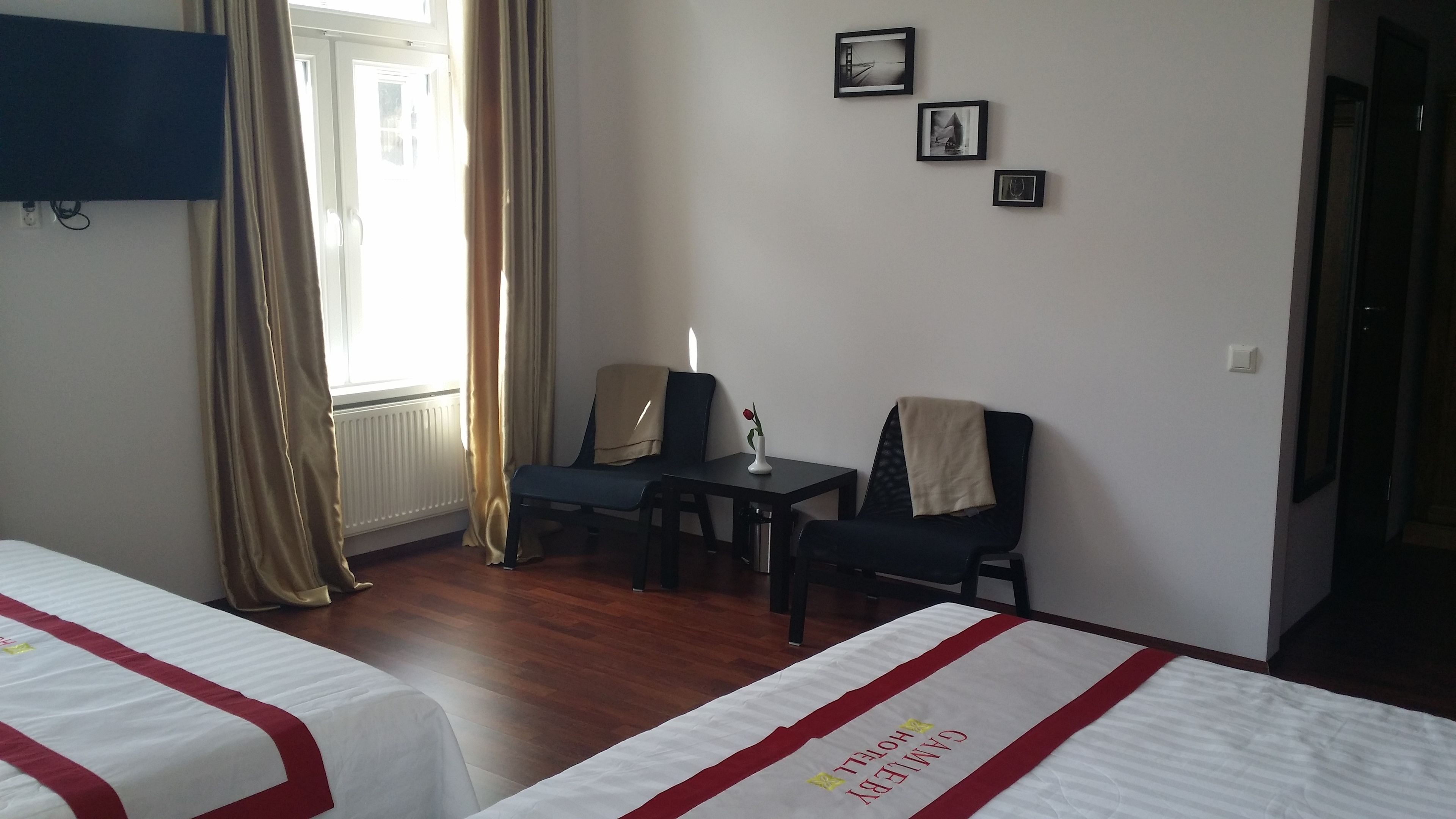 Quarto de hotel com duas camas, uma mesinha, duas cadeiras, um vaso com uma flor e três quadros na parede.