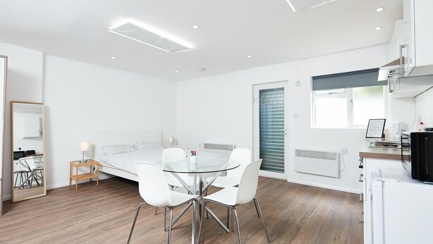 Quarto moderno e bem iluminado, com mesa de vidro redonda, cadeiras brancas, cama, pequena mesa de cabeceira de madeira com abajur e um espaço de cozinha compacta.