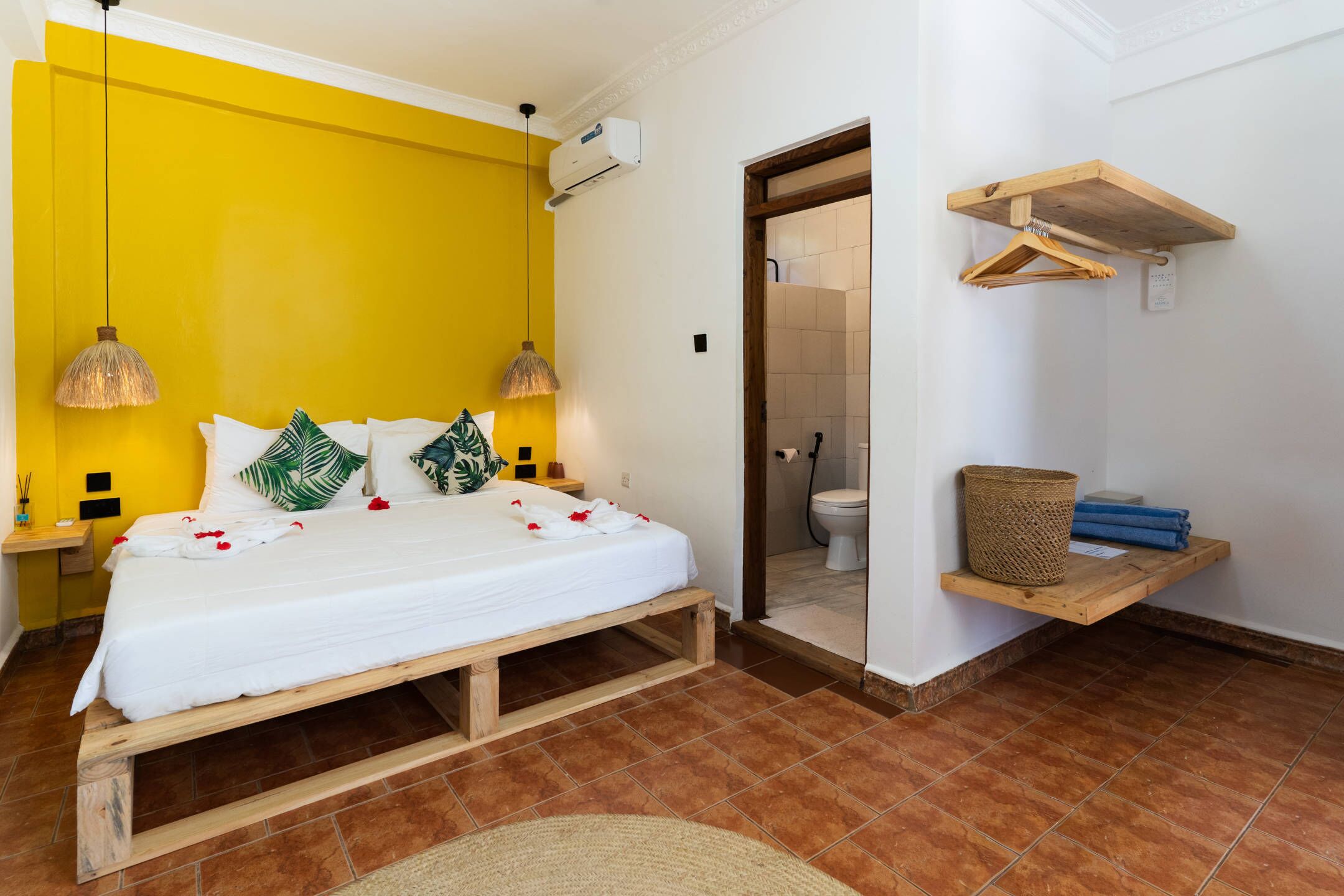 Quarto com cama de madeira, parede amarela de destaque, prateleira de madeira com cesta e luminária pendente.