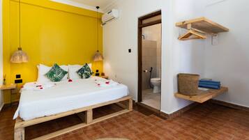 Quarto com cama de madeira, parede amarela de destaque, prateleira de madeira com cesta e luminária pendente.