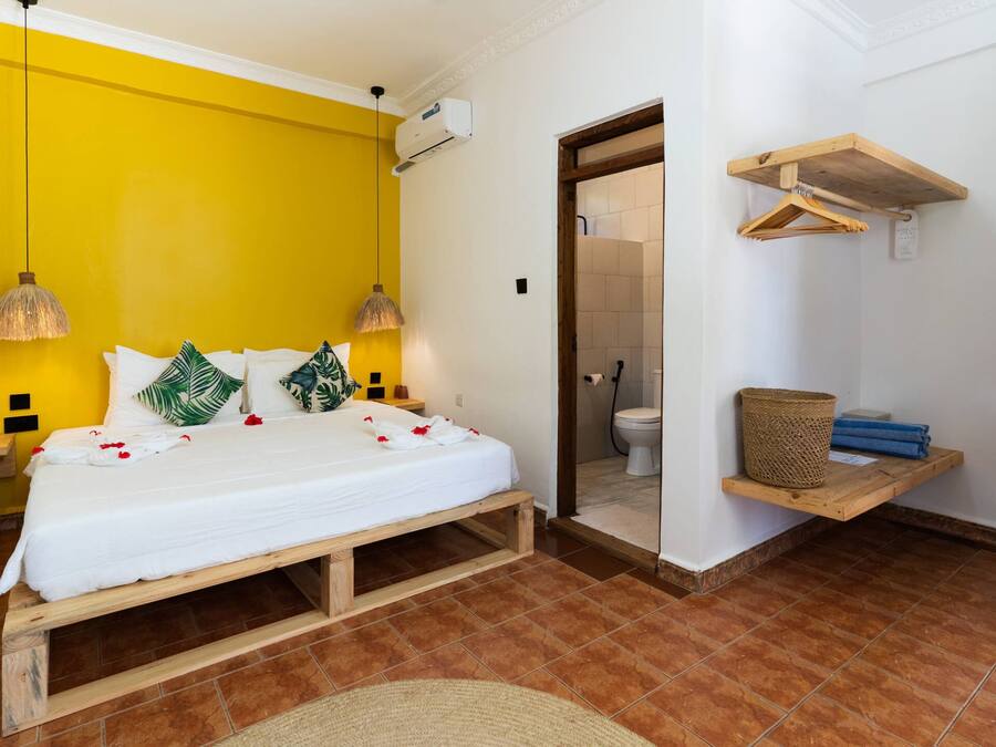 Quarto com cama de madeira, parede amarela de destaque, prateleira de madeira com cesta e luminária pendente.