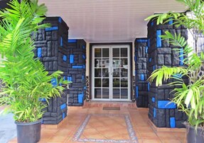 Entrada de um edifício com piso de azulejos, porta de vidro e duas plantas em vasos.