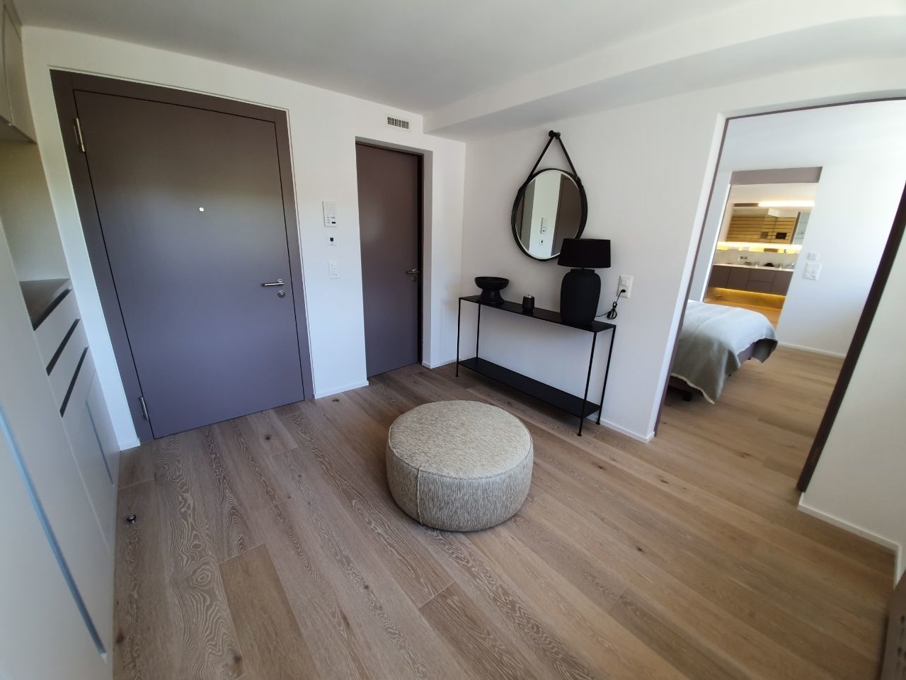 Quarto de hotel moderno com piso de madeira, um banquinho redondo, um espelho e uma cama ao fundo.