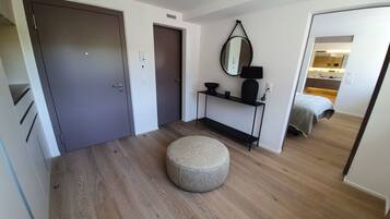 Quarto de hotel moderno com piso de madeira, um banquinho redondo, um espelho e uma cama ao fundo.