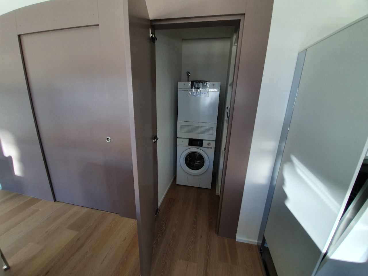 Uma lavanderia compacta com máquina de lavar e secadora, piso de madeira e uma porta que dá para outro cômodo.