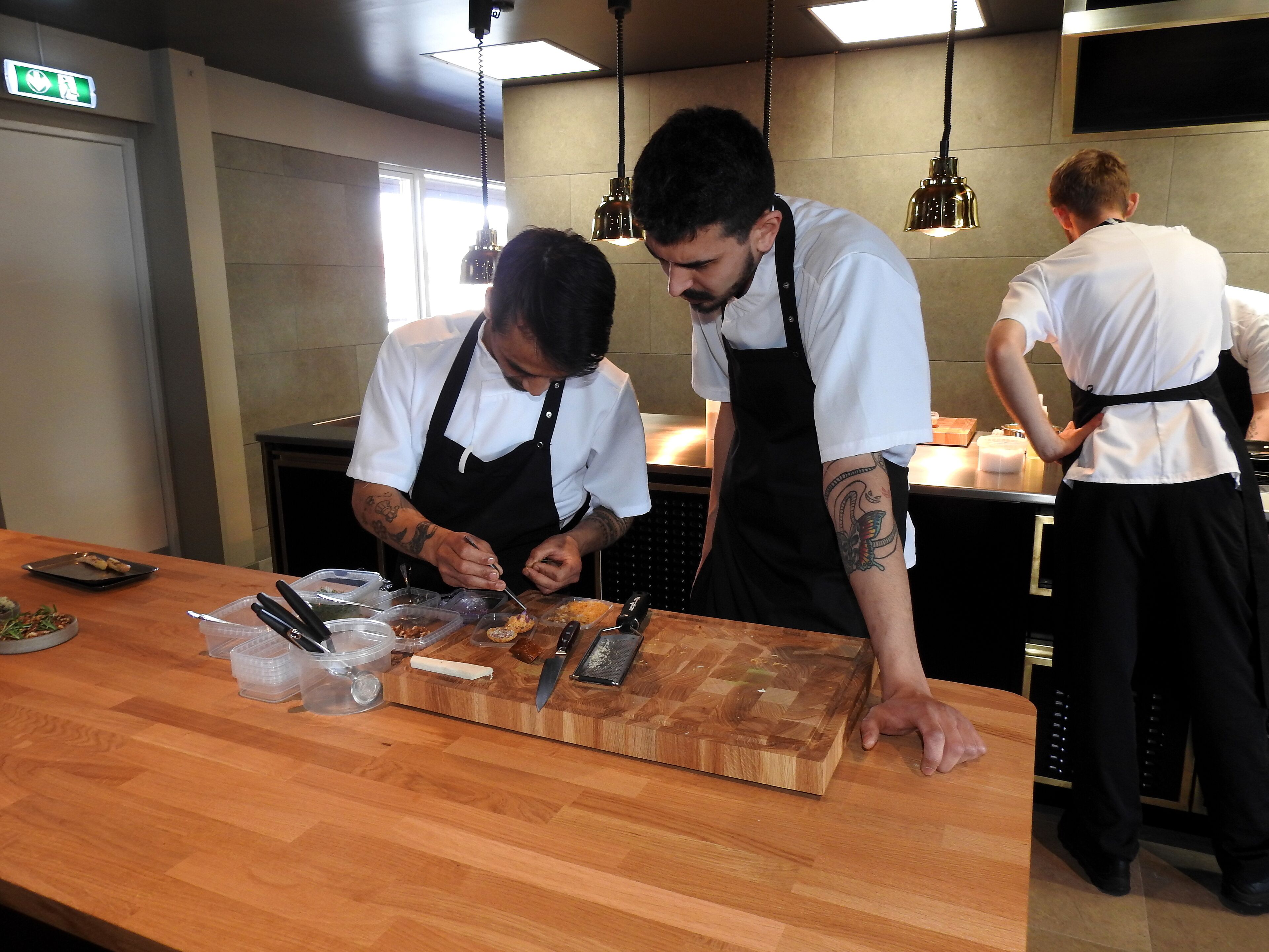 Cozinha com chefs preparando alimentos, bancada de madeira e diversos utensílios culinários.