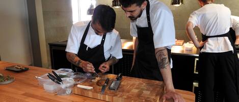 Cozinha com chefs preparando alimentos, bancada de madeira e diversos utensílios culinários.