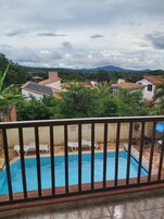 Piscina com deck ladrilhado, rodeada por uma cerca de madeira. Ao fundo, casas com telhados de barro avermelhado e vegetação exuberante.